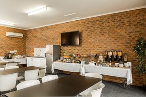 Sala de conferência com parede de tijolos, mesas e cadeiras brancas, equipada com frigorífico, espaço para café e uma TV de tela plana.