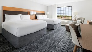 Quarto de hotel com duas camas, uma mesa, uma cadeira e uma janela grande com vista.