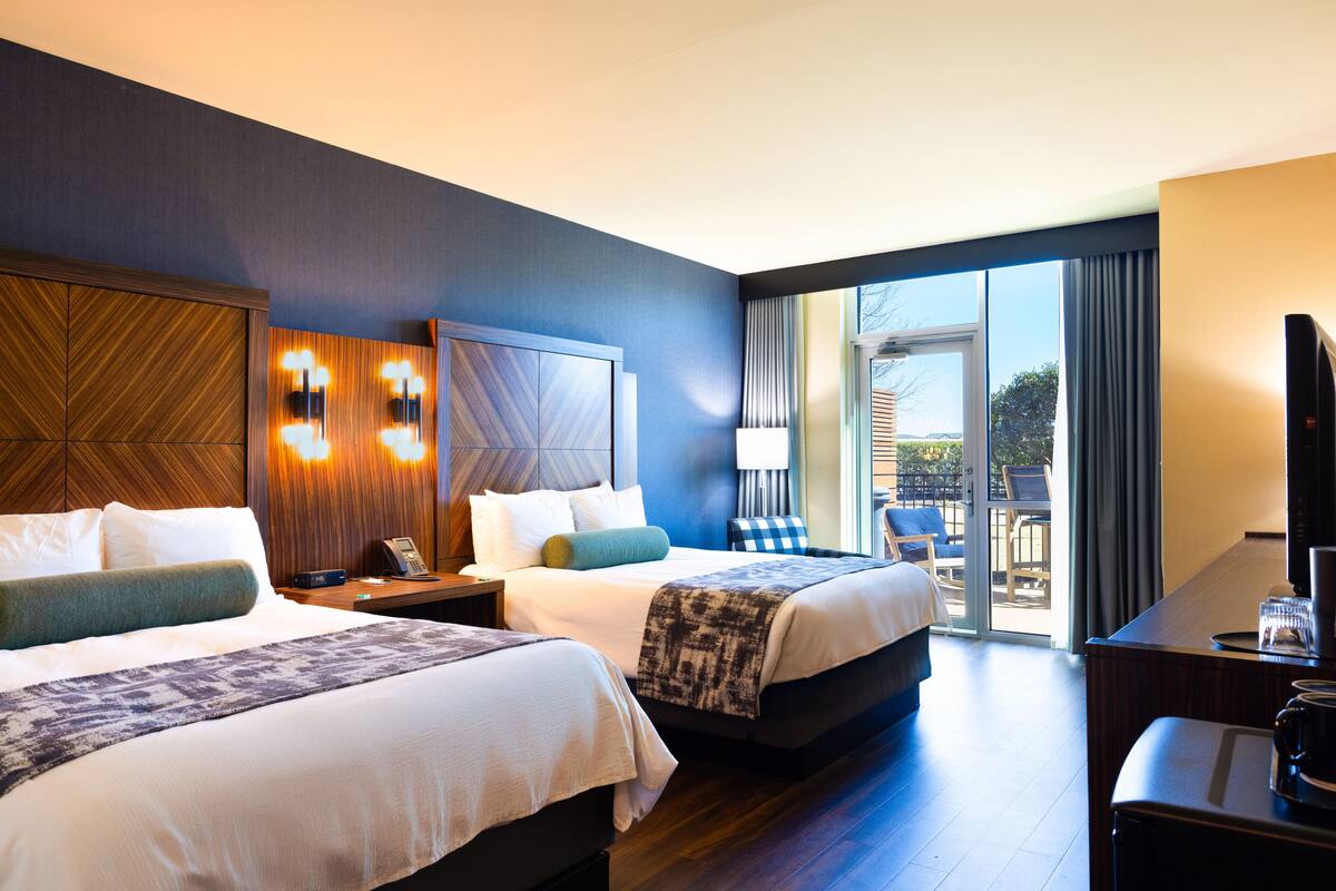 Quarto de hotel com duas camas, uma escrivaninha, televisão e varanda com área de estar ao ar livre.