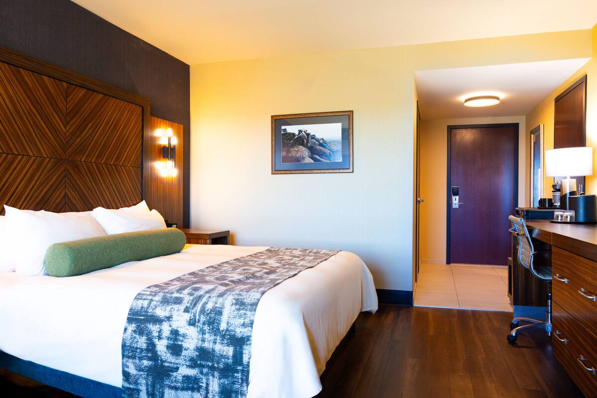 Quarto de hotel com uma cama grande, uma escrivaninha com abajur, um aparador e um quadro na parede.