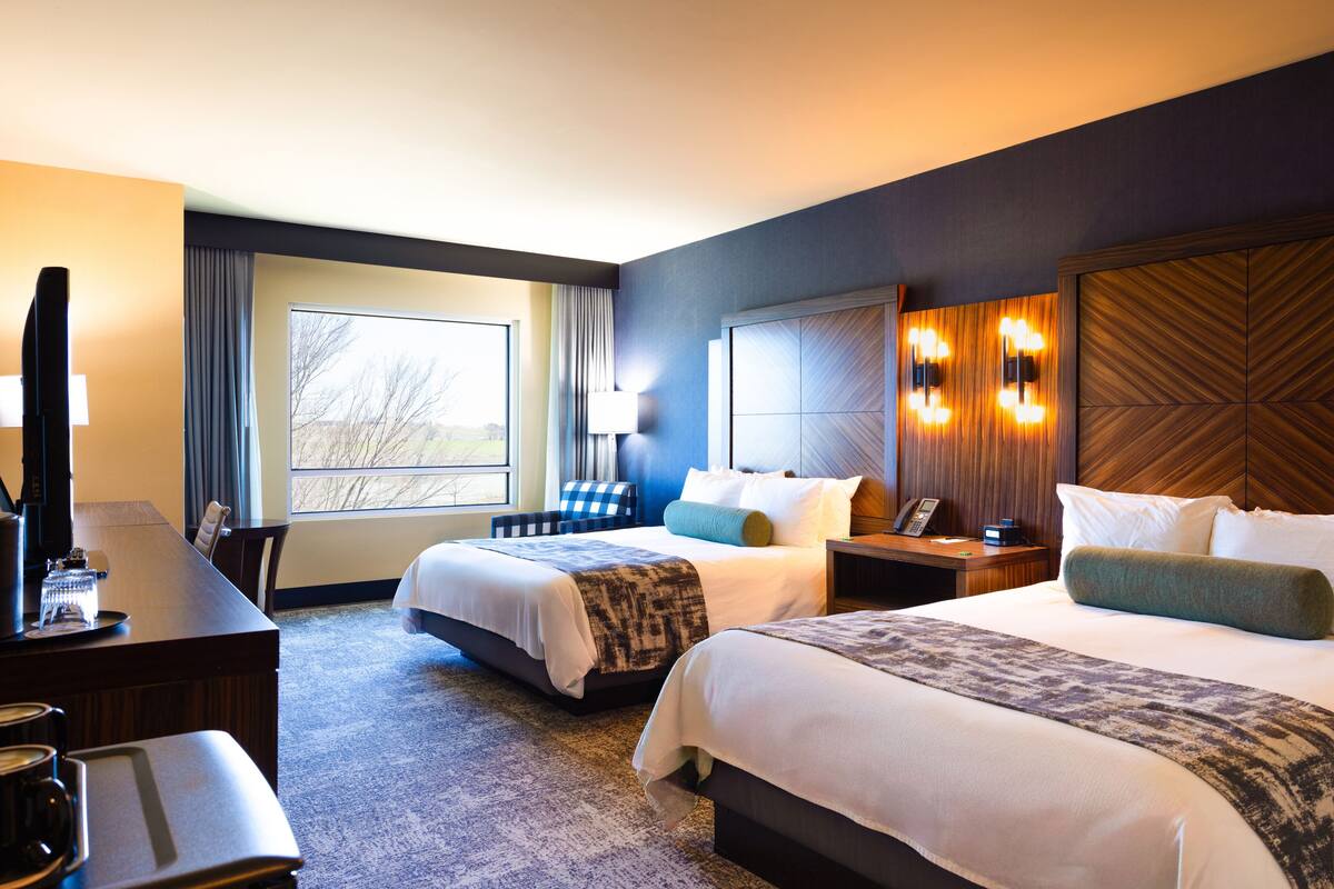 Quarto de hotel com duas camas, uma escrivaninha, televisão e janela com cortinas.
