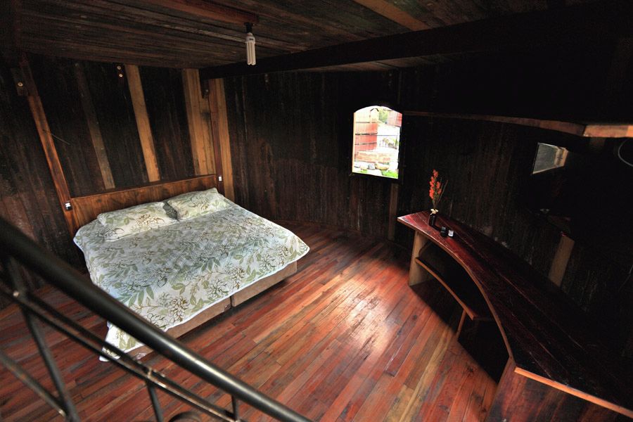 Um quarto rústico com piso de madeira, uma cama com roupa de cama floral, uma mesa pequena e uma janela com vista.