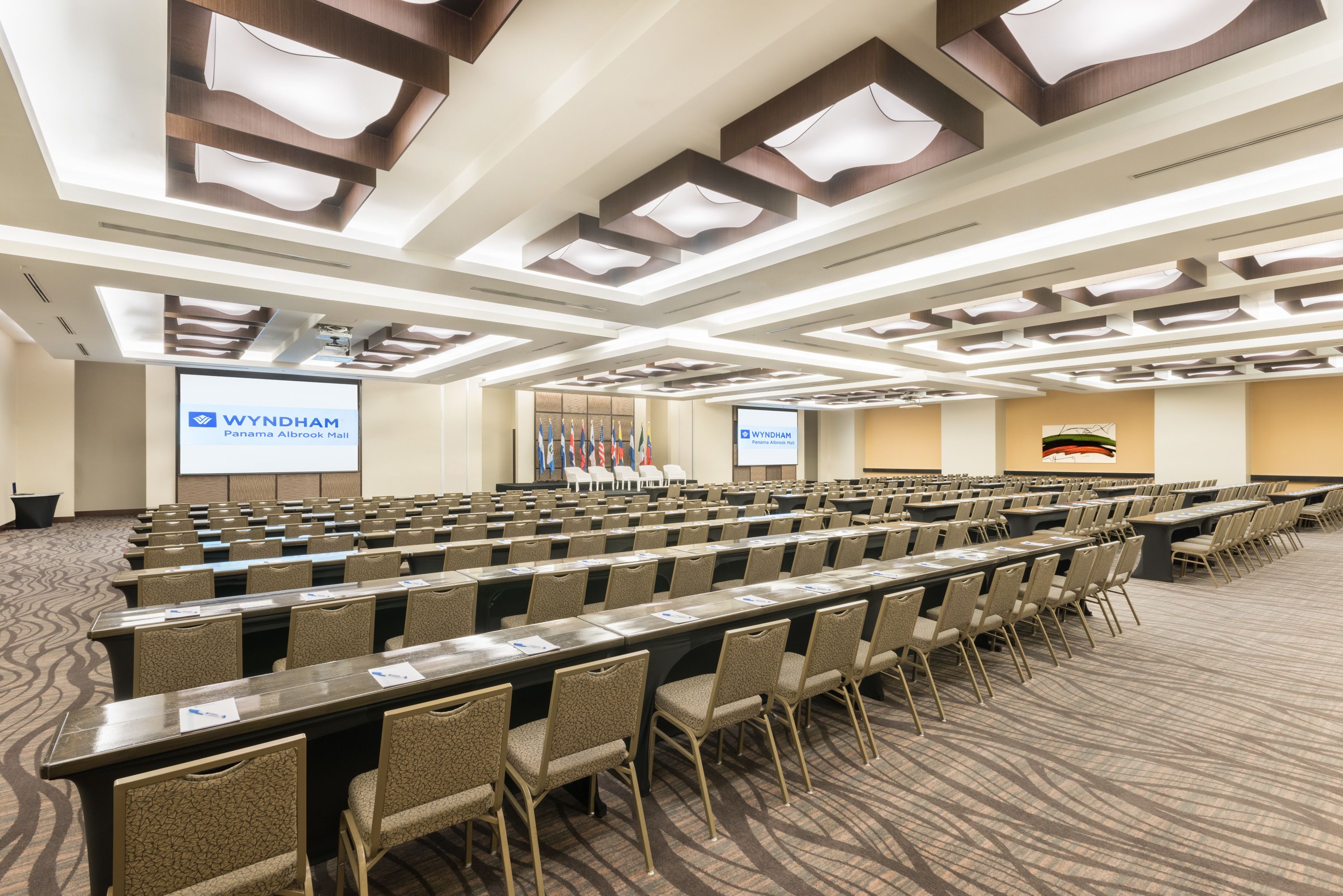 Uma sala de conferências com fileiras de mesas e cadeiras, uma tela grande e uma área para apresentações.