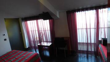 Quarto de hotel com uma cama, uma escrivaninha e duas janelas com cortinas leves.