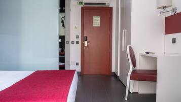 Quarto de hotel com uma cama, uma escrivaninha, uma cadeira, um abajur e uma porta com sinalização de saída de emergência.