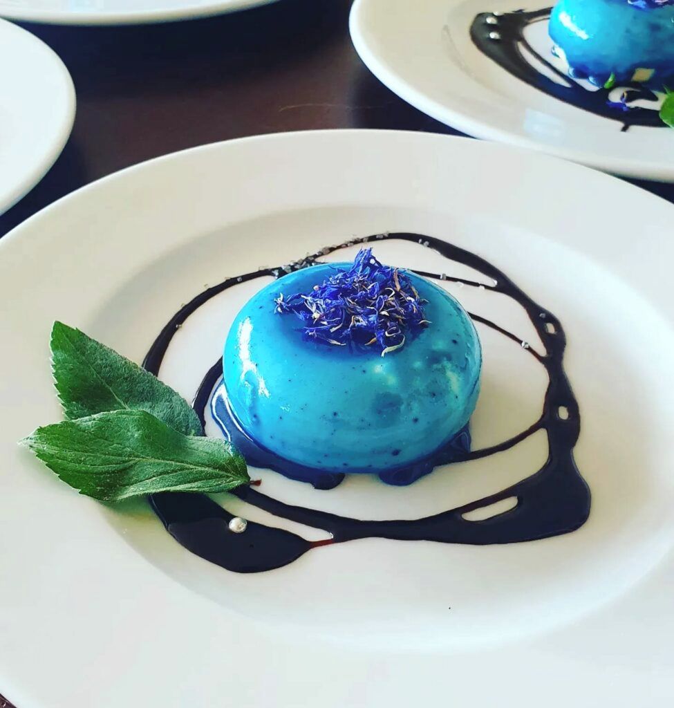 Sobremesa em um prato branco com cobertura azul, decorada com folhas de hortelã e um fio de molho de chocolate.