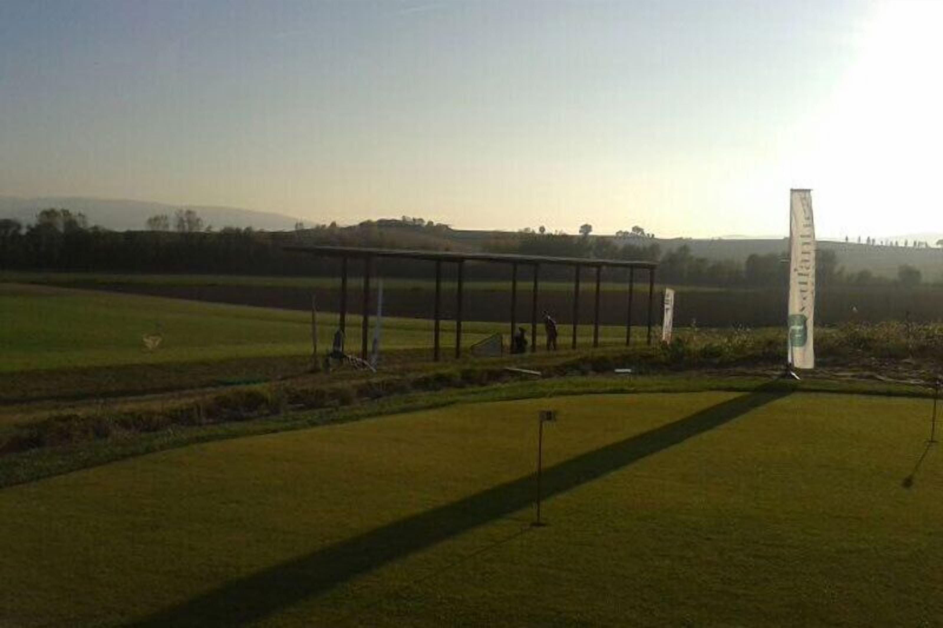 Campo de golfe com driving range coberto e haste de bandeira em primeiro plano.