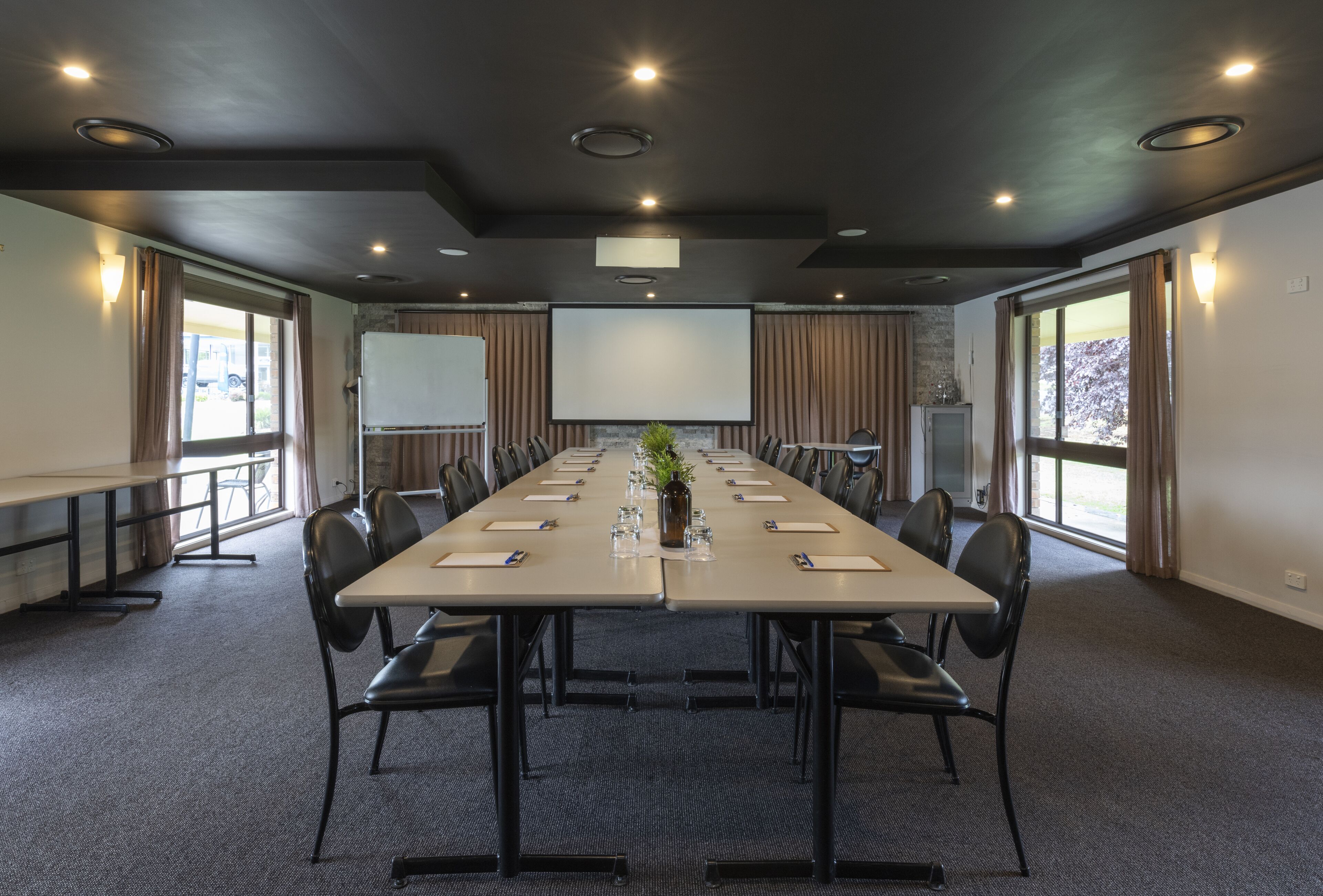 Sala de conferências com uma mesa comprida, cadeiras, tela de projetor e uma janela grande.