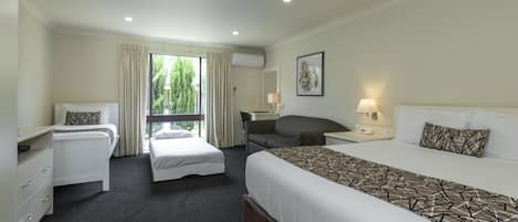 Quarto de hotel com uma cama grande, uma cama menor, um sofá, uma escrivaninha e uma cadeira. Há uma janela com cortinas, um abajur e um quadro na parede.