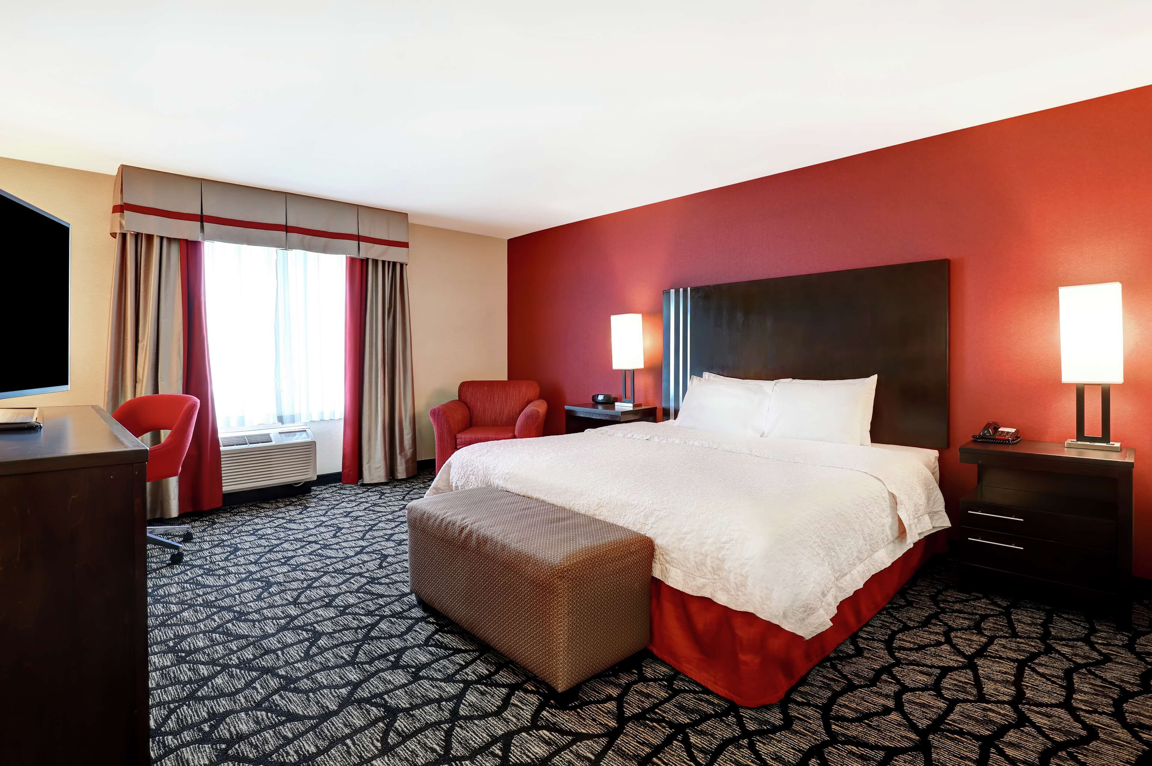 Quarto de hotel com uma cama grande, uma escrivaninha, uma cadeira, uma televisão e uma janela com cortinas.