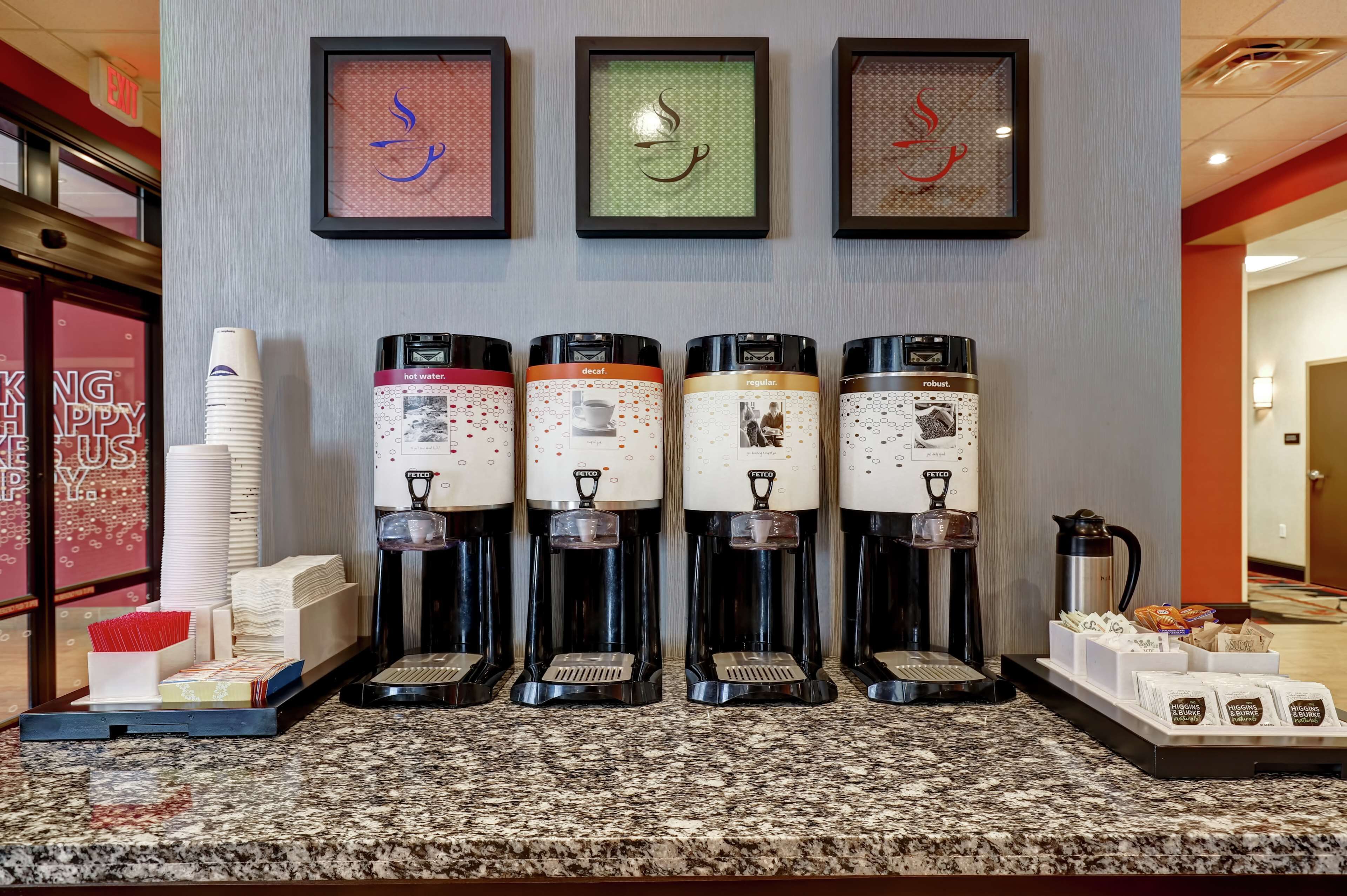 Um espaço de café com quatro dispensadores, uma pilha de copos descartáveis e uma bandeja com cápsulas de café.