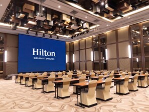 Uma sala de conferências com uma tela grande exibindo 'Hilton Sukhumvit Bangkok' e fileiras de mesas com cadeiras.