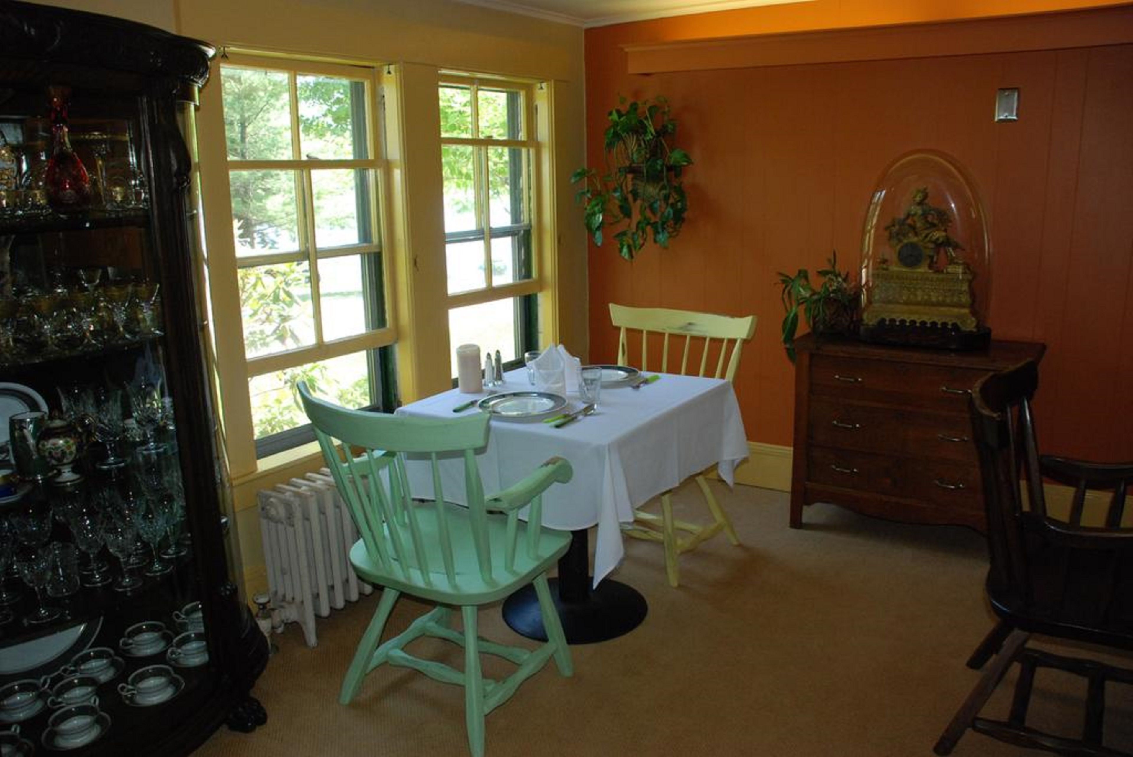 Uma área de refeições com uma mesa posta para dois, uma cadeira verde, um armário de madeira e uma janela com vista para vegetação.