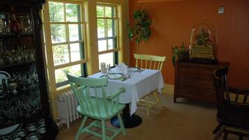 Uma área de refeições com uma mesa posta para dois, uma cadeira verde, um armário de madeira e uma janela com vista para vegetação.