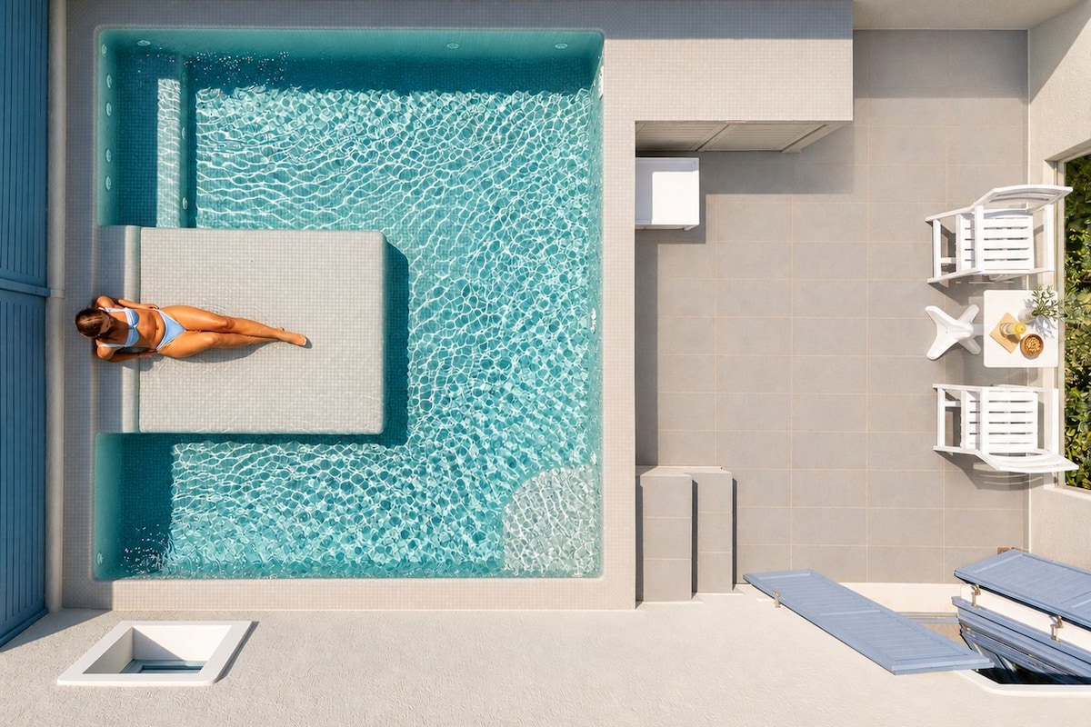 Uma área moderna de piscina ao ar livre, com uma pessoa relaxando em uma espreguiçadeira, um espaço de estar pequeno com mesa e cadeiras, e uma área coberta com vista para a piscina.