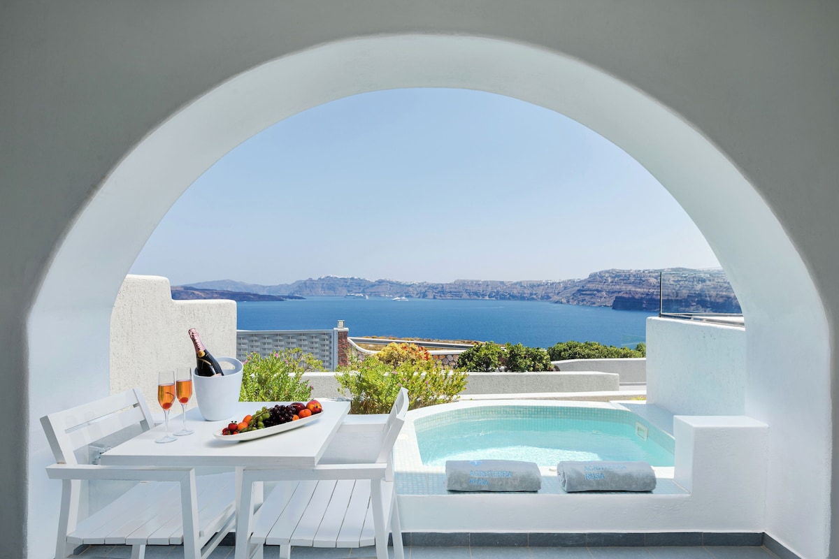 Terraço banhado por luz solar, com piscina, móveis brancos e mesa posta com champanhe, taças e frutas.