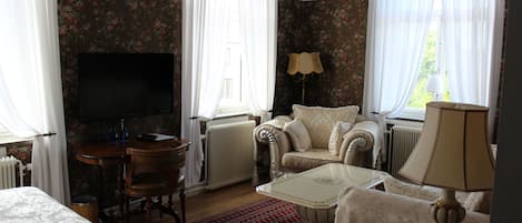 Quarto de hotel com cama, televisão, sofá, mesinha, candeeiro e janela com cortinas.