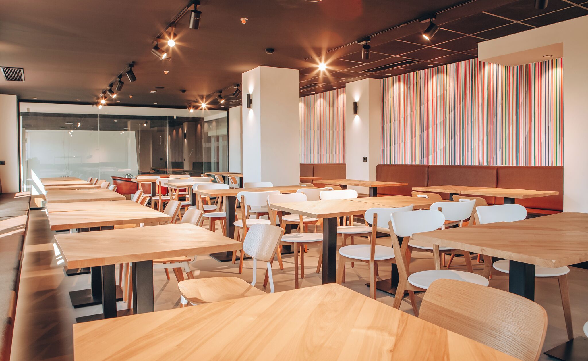 Interior moderno de restaurante com mesas e cadeiras de madeira, decoração na parede com listras e iluminação embutida no teto.