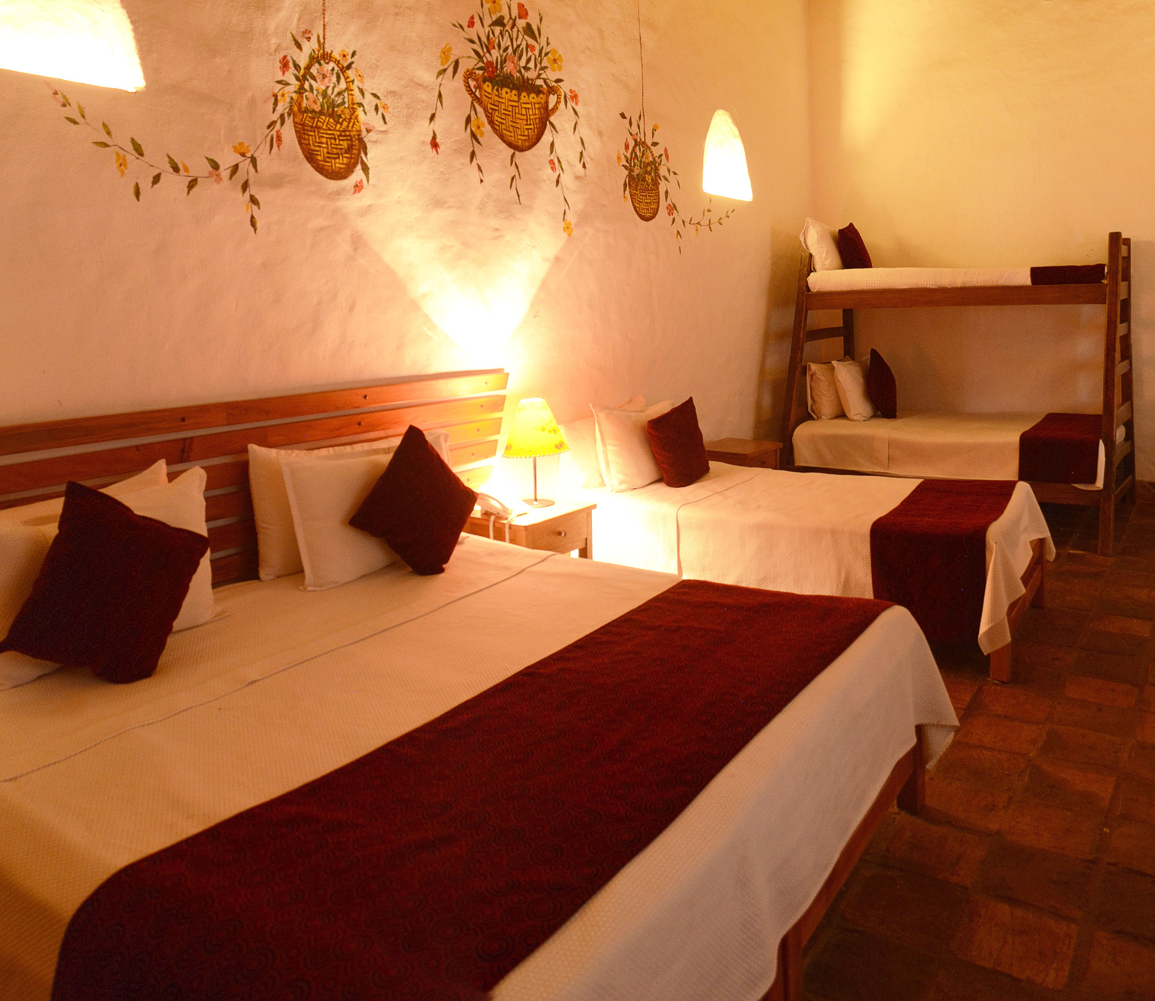 Quarto de hotel com duas camas, cada uma com roupa de cama branca e mantas vermelhas. Há cestos suspensos com flores na parede, e uma escada de madeira que leva a uma beliche superior.