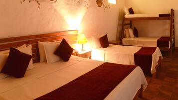 Quarto de hotel com duas camas, cada uma com roupa de cama branca e mantas vermelhas. Há cestos suspensos com flores na parede, e uma escada de madeira que leva a uma beliche superior.