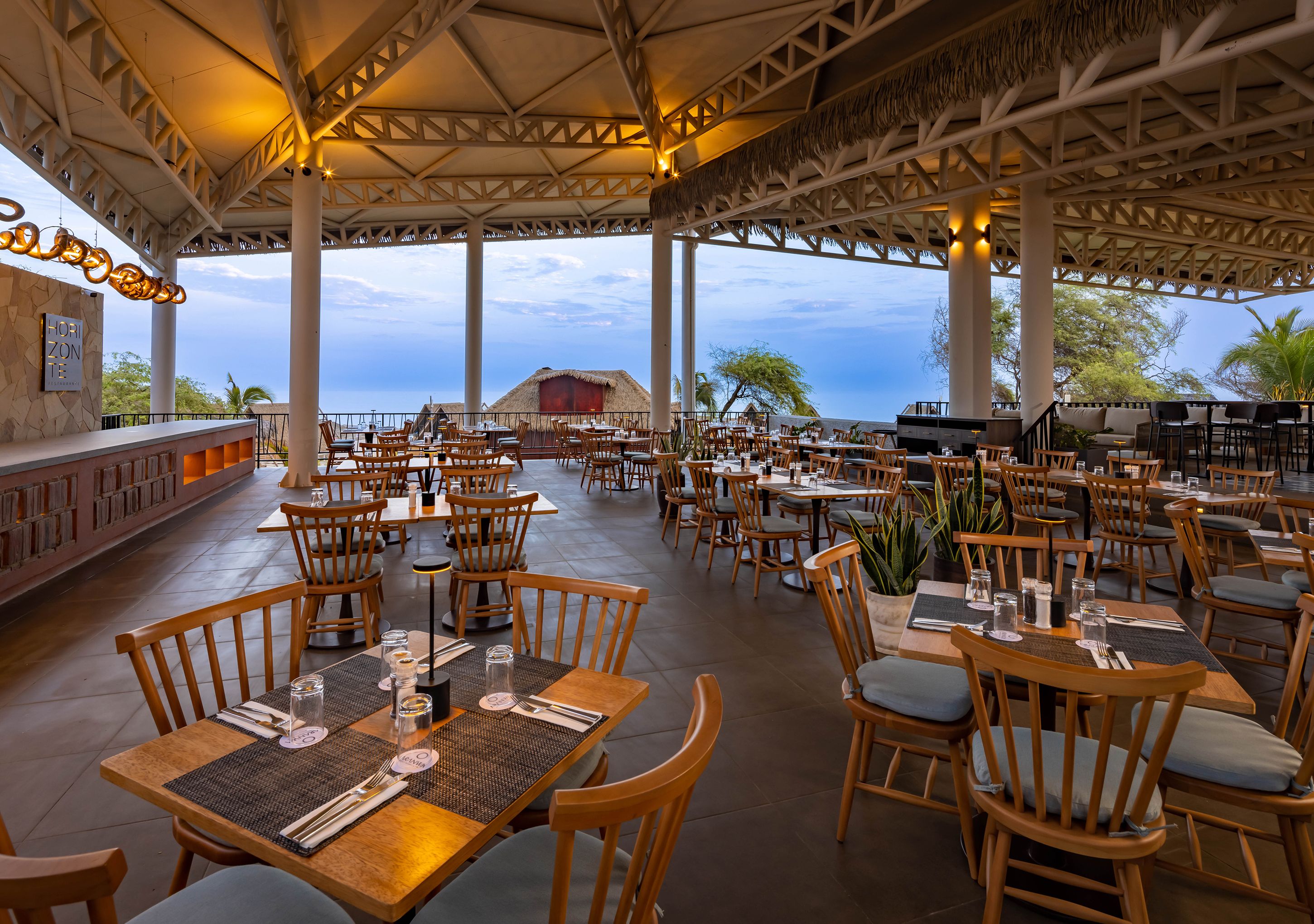 Restaurante com mesas e cadeiras de madeira e estrutura com telhado de palha, oferecendo vista para o oceano.