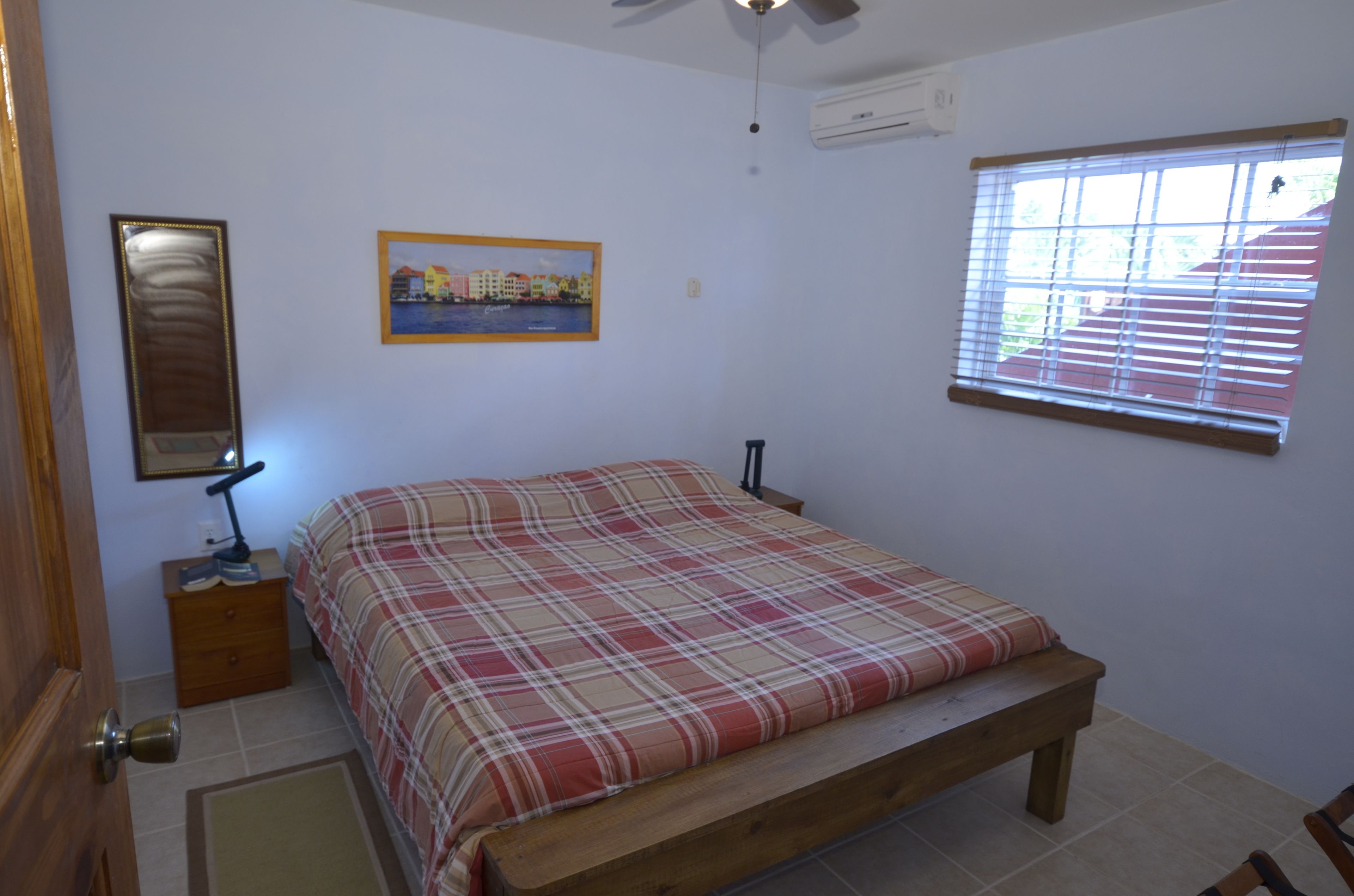 Quarto com cama, mesa de cabeceira, espelho, pintura e janela com persianas.