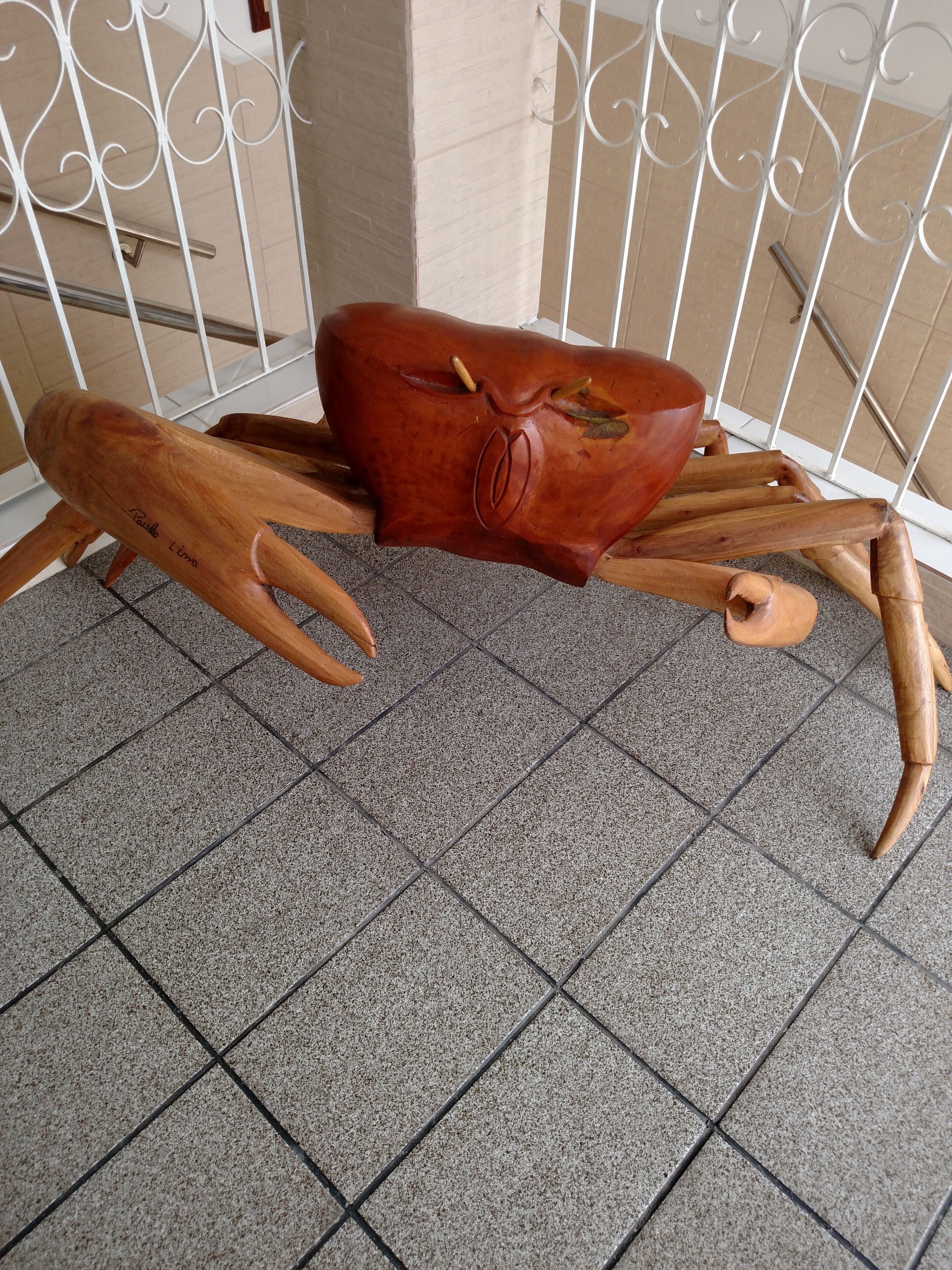 Uma escultura de madeira que se assemelha a um caranguejo, com uma sacola castanha colocada em cima, sobre um chão de azulejos e com um corrimão de metal ao fundo.