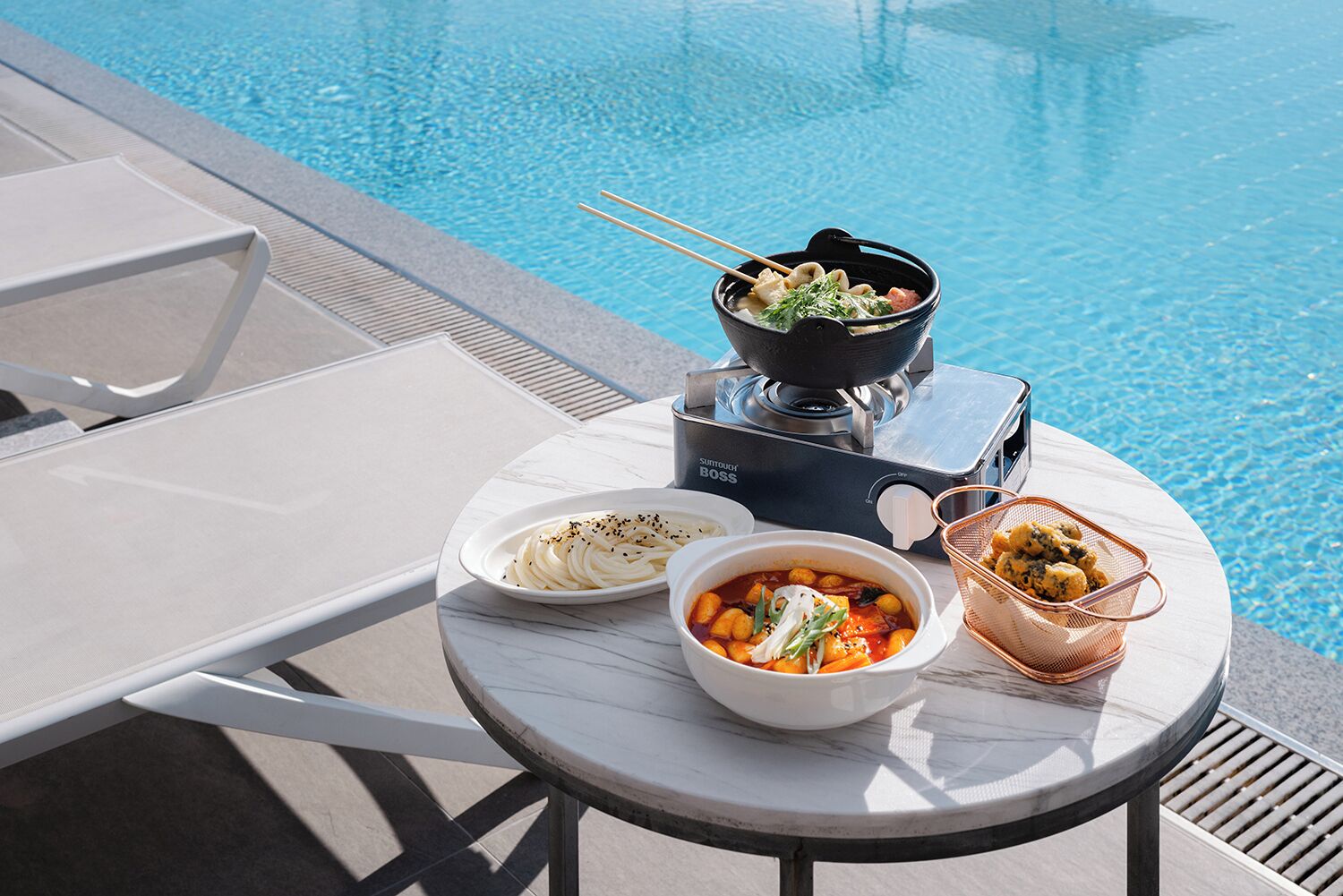 Uma mesa de jantar com um fogão portátil, tigelas de comida e uma cadeira à beira da piscina.
