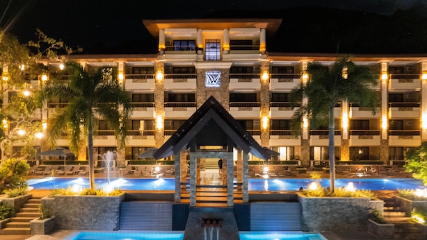 Um hotel com vários andares, piscina, palmeiras e entrada bem iluminada.