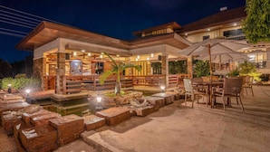 Uma área de pátio externa com assentos de pedra, mesas e guarda-sóis, cercada por uma piscina e um edifício com área de estar coberta.