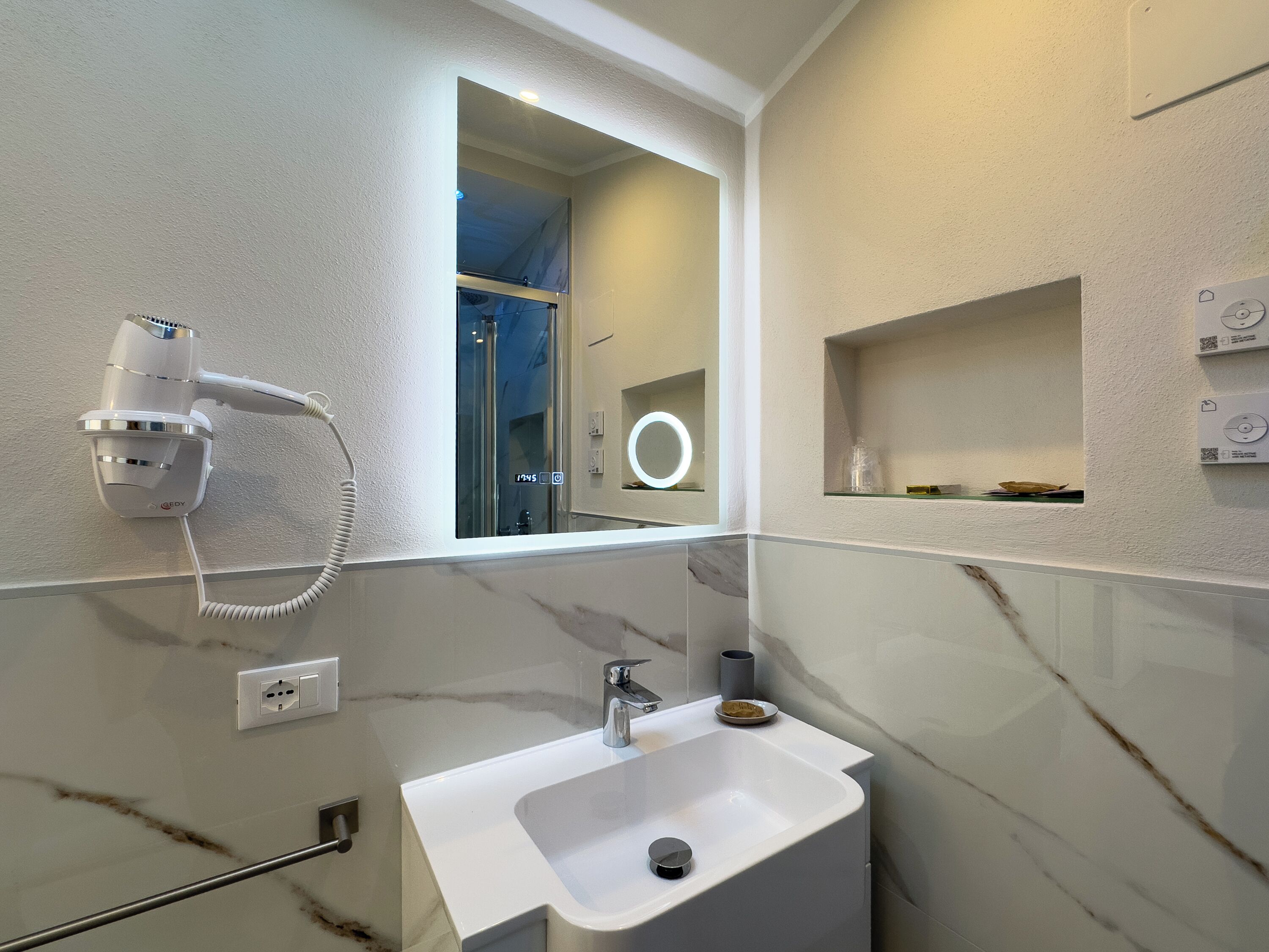 Casa de banho moderna com lavatório branco, secador de cabelo fixado na parede, espelho com luz integrada e uma pequena prateleira com artigos de higiene pessoal.