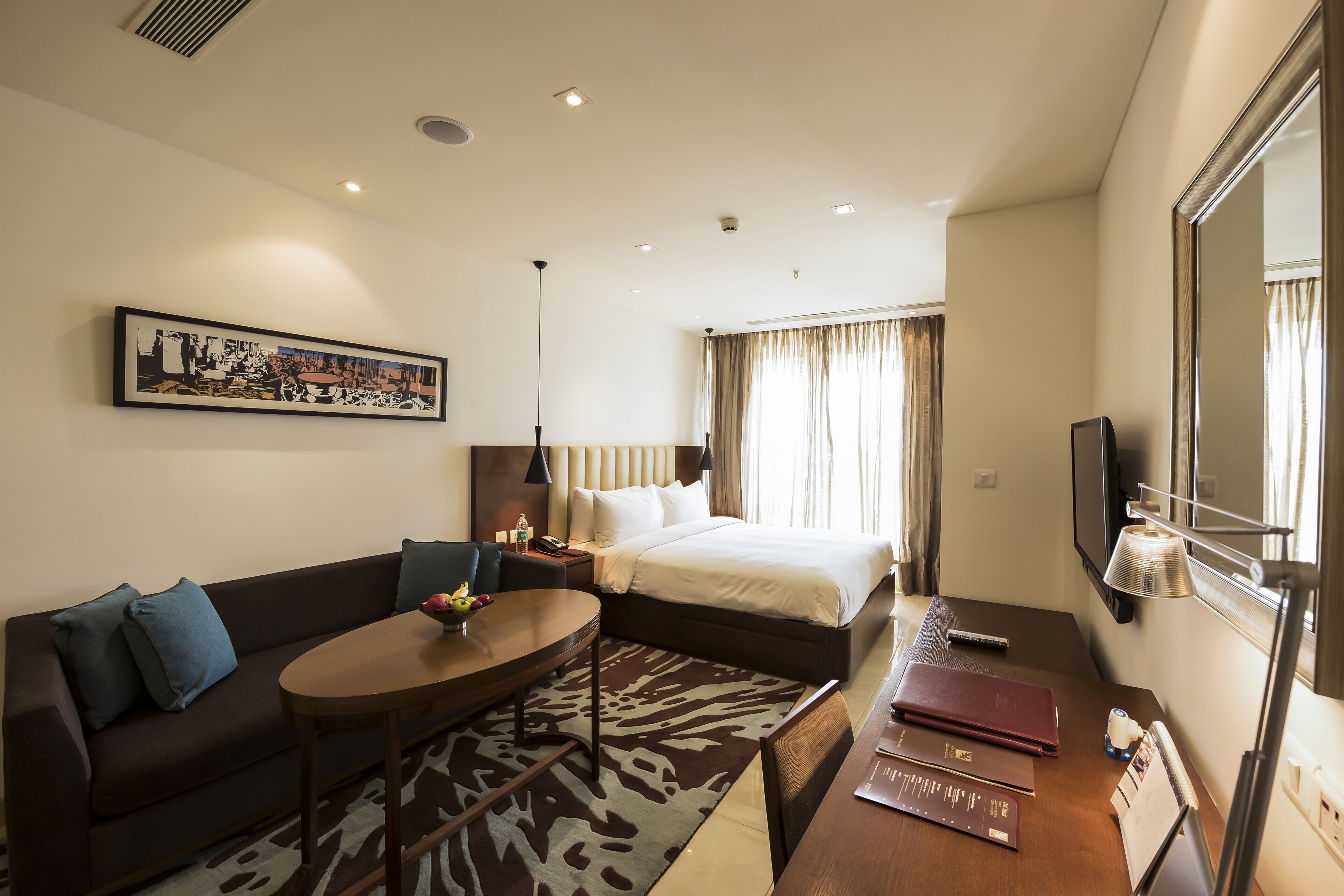 Quarto de hotel com cama, sofá, escrivaninha, cadeira, luminária, televisão e um quadro na parede.