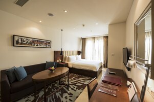 Quarto de hotel com cama, sofá, escrivaninha, cadeira, luminária, televisão e um quadro na parede.