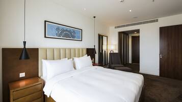 Quarto de hotel com uma cama grande, mesa de cabeceira, abajur, cadeira e porta.