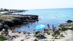 Uma praia rochosa com água azul cristalina, pessoas nadando e tomando sol, e alguns guarda-sóis oferecendo sombra.