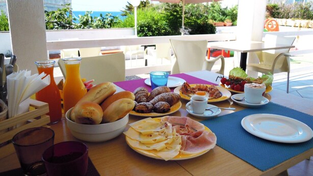 Mesa de café da manhã com croissants, doces, ovos e presunto, com vista para o mar e vegetação ao fundo.