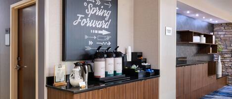 Uma área de recepção de hotel com uma placa preta com a inscrição "Spring Forward", um canto com café e uma parede de pedra.