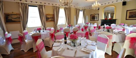 Salão de banquetes com mesas redondas, toalhas brancas e cadeiras adornadas com fitas cor-de-rosa.
