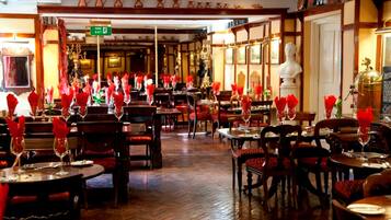 Restaurante com piso de madeira, cadeiras estofadas em vermelho e mesas preparadas com guardanapos e copos de vinho vermelhos.