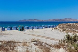 Uma praia com tendas e uma pequena estrutura, provavelmente um banheiro, com vista clara para o mar e colinas ao longe.