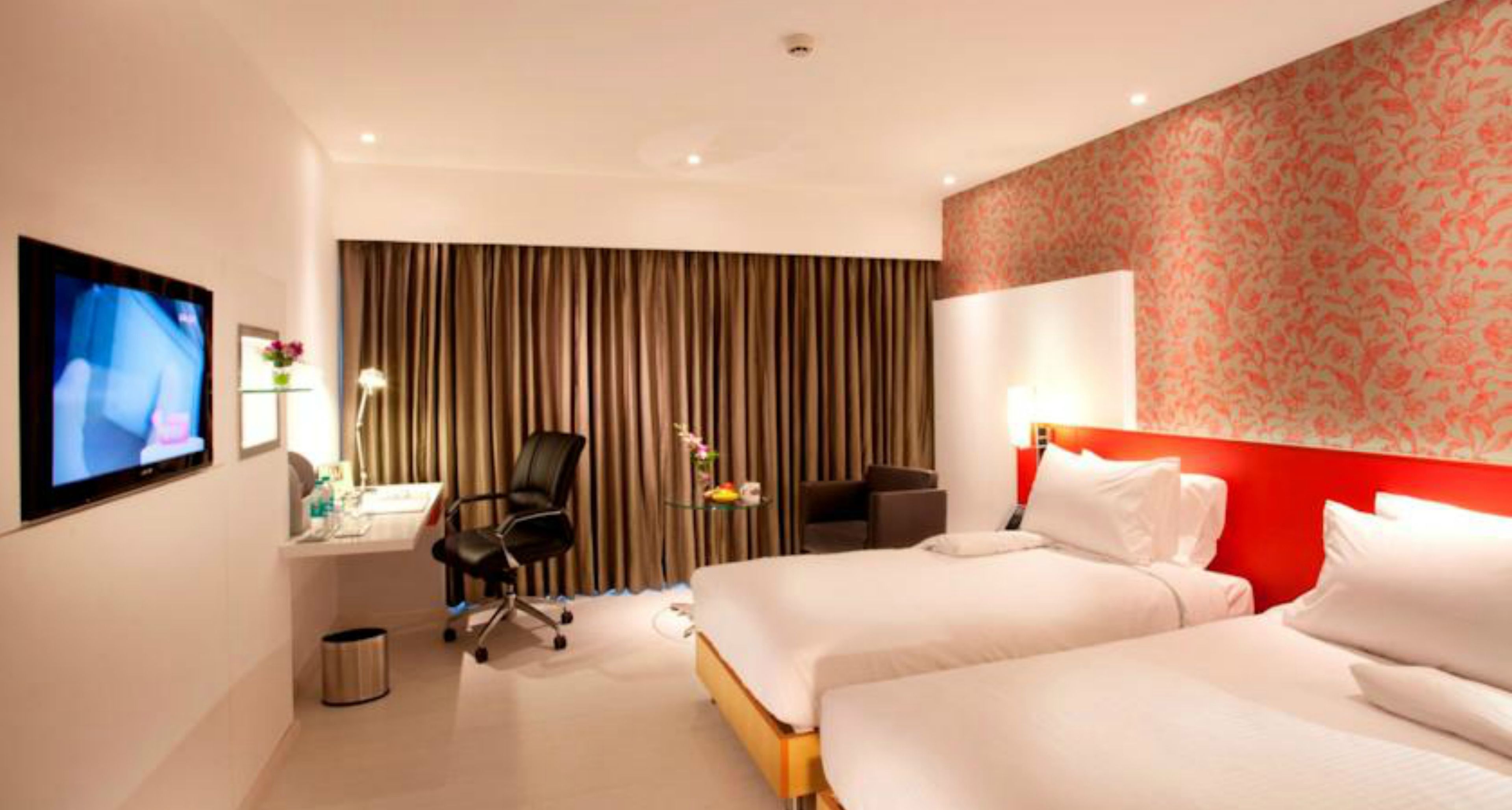 Quarto de hotel com duas camas, uma mesa com cadeira, televisão e uma janela grande com cortinas.