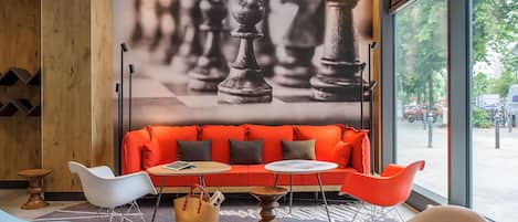 Uma área de estar moderna com um sofá vermelho, cadeiras brancas e laranja, uma mesa redonda e uma obra de arte com um tabuleiro de xadrez.