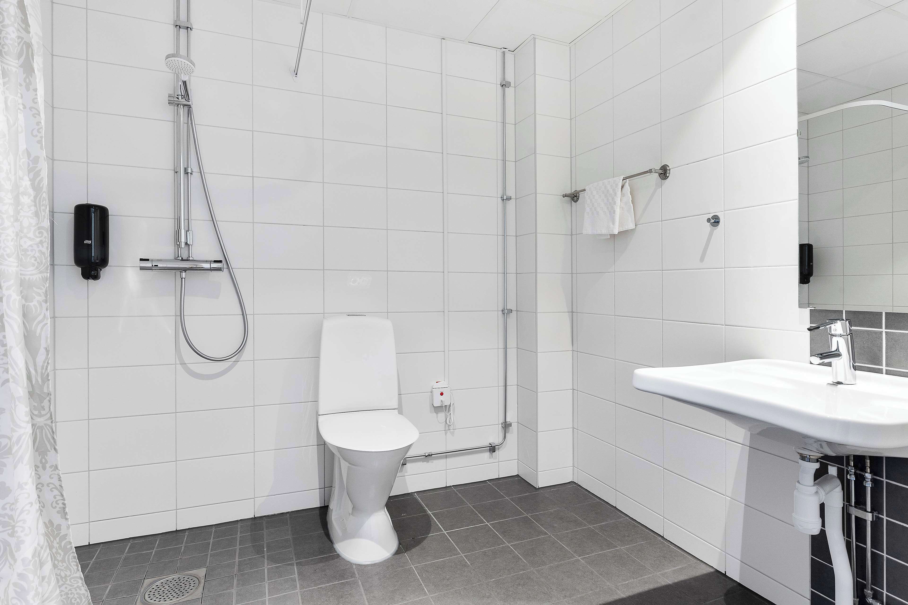 Casa de banho moderna com sanita branca, chuveiro com chuveiro manual, lavatório com espelho e um porta-toalhas.