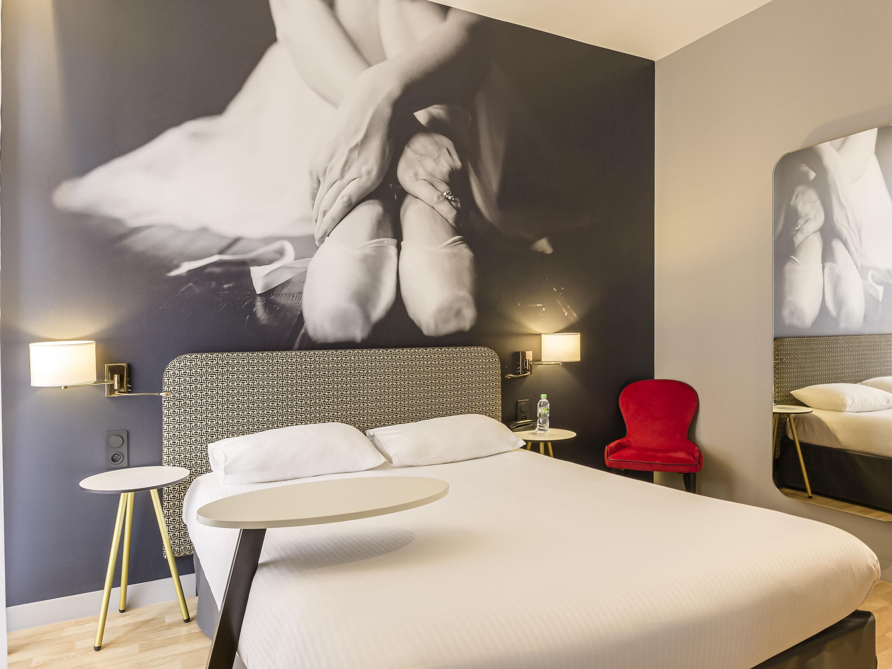 Quarto de hotel com uma cama grande, uma cadeira vermelha, duas mesinhas e luminárias fixadas na parede.