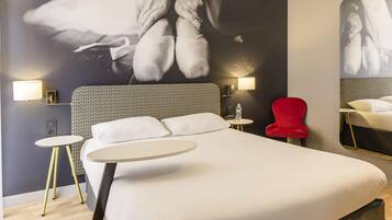 Quarto de hotel com uma cama grande, uma cadeira vermelha, duas mesinhas e luminárias fixadas na parede.