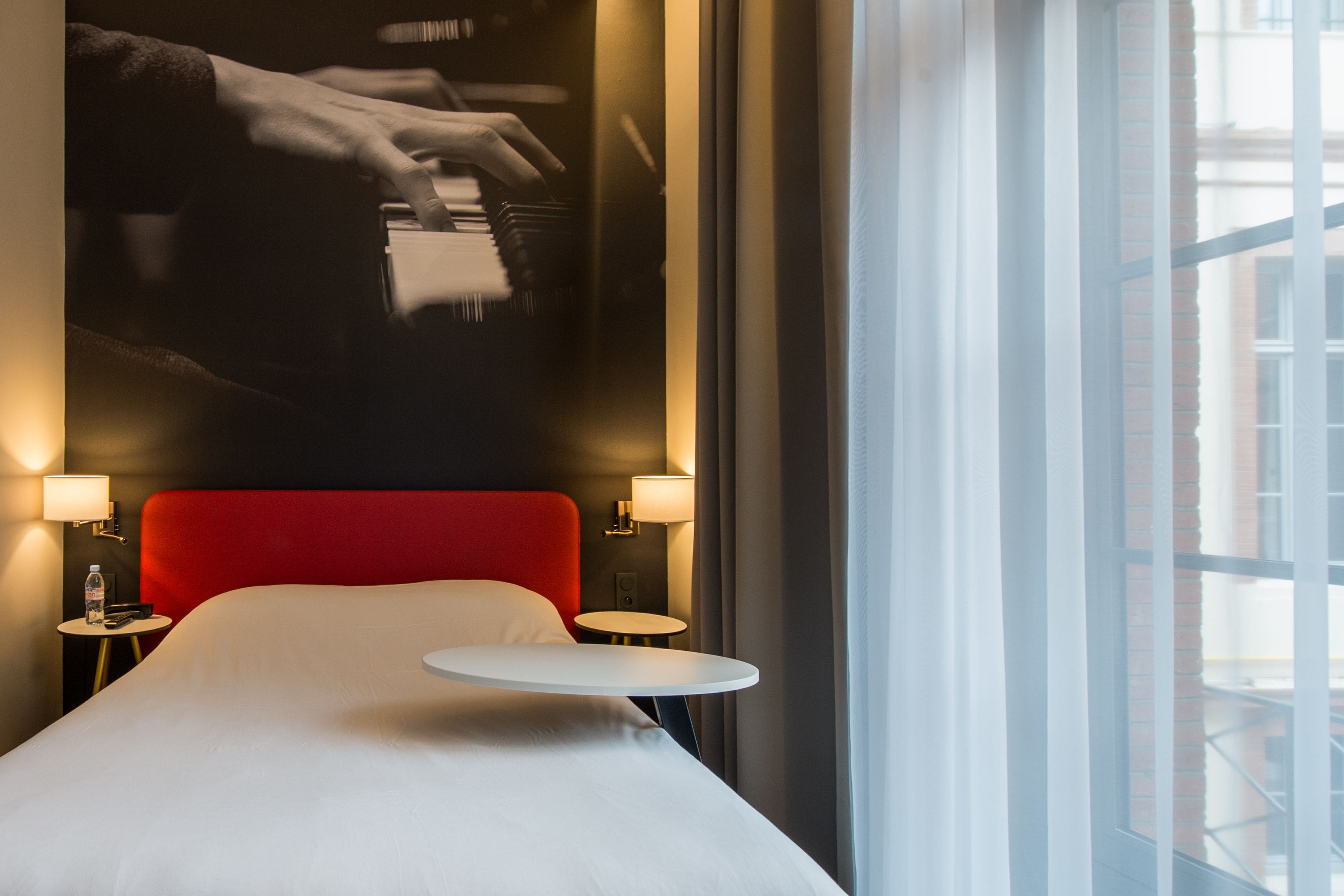 Quarto de hotel com cabeceira vermelha, cama branca, mesa redonda e um cartaz com mãos tocando piano.