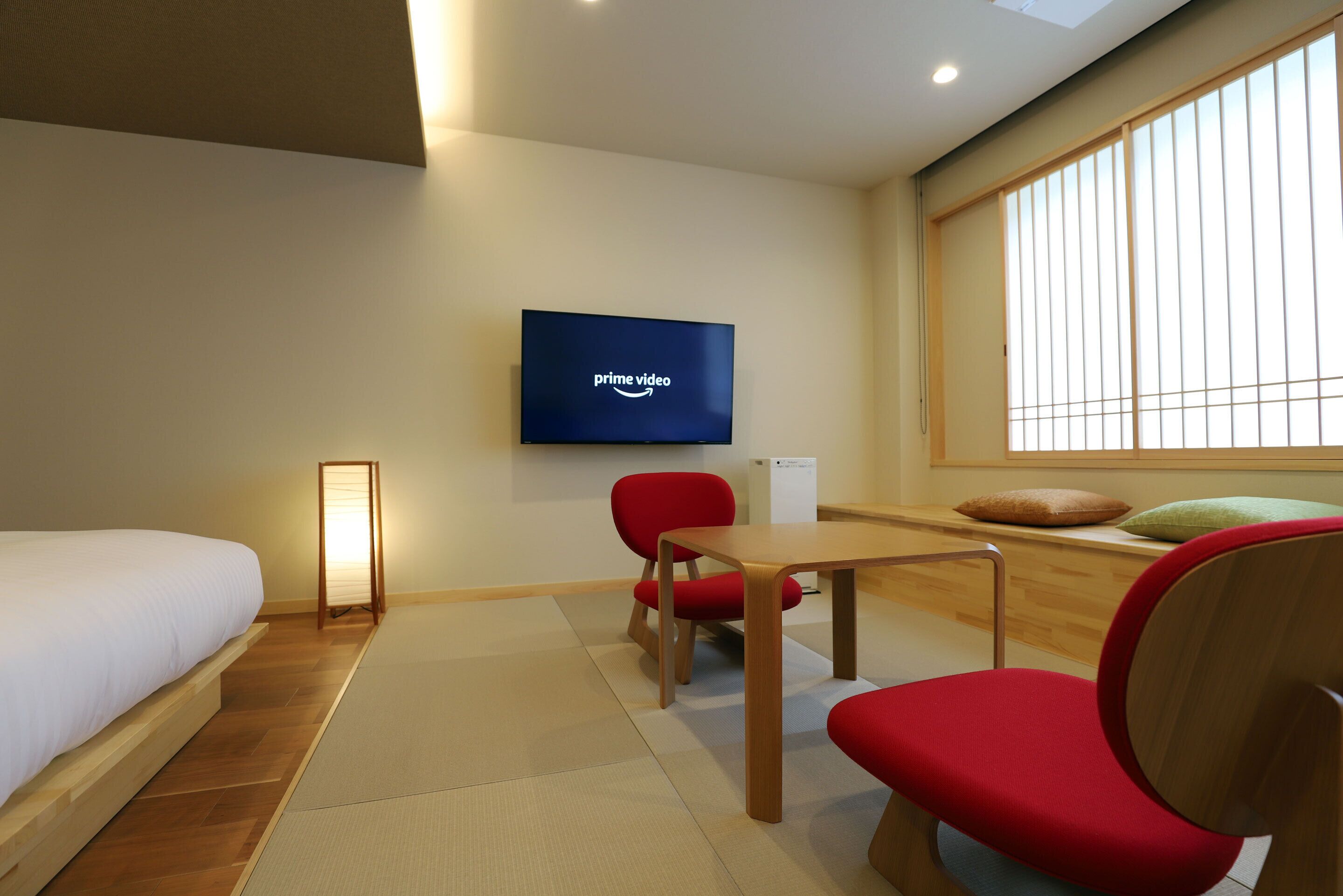 Quarto de hotel com cama, televisão fixada na parede, mesa, duas cadeiras vermelhas e janela com persianas.