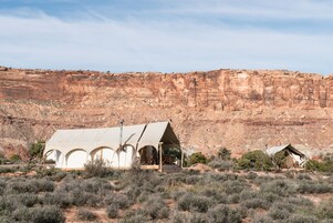 Um acampamento no deserto com uma tenda grande e estruturas menores, rodeado por vegetação rala e um penhasco rochoso.