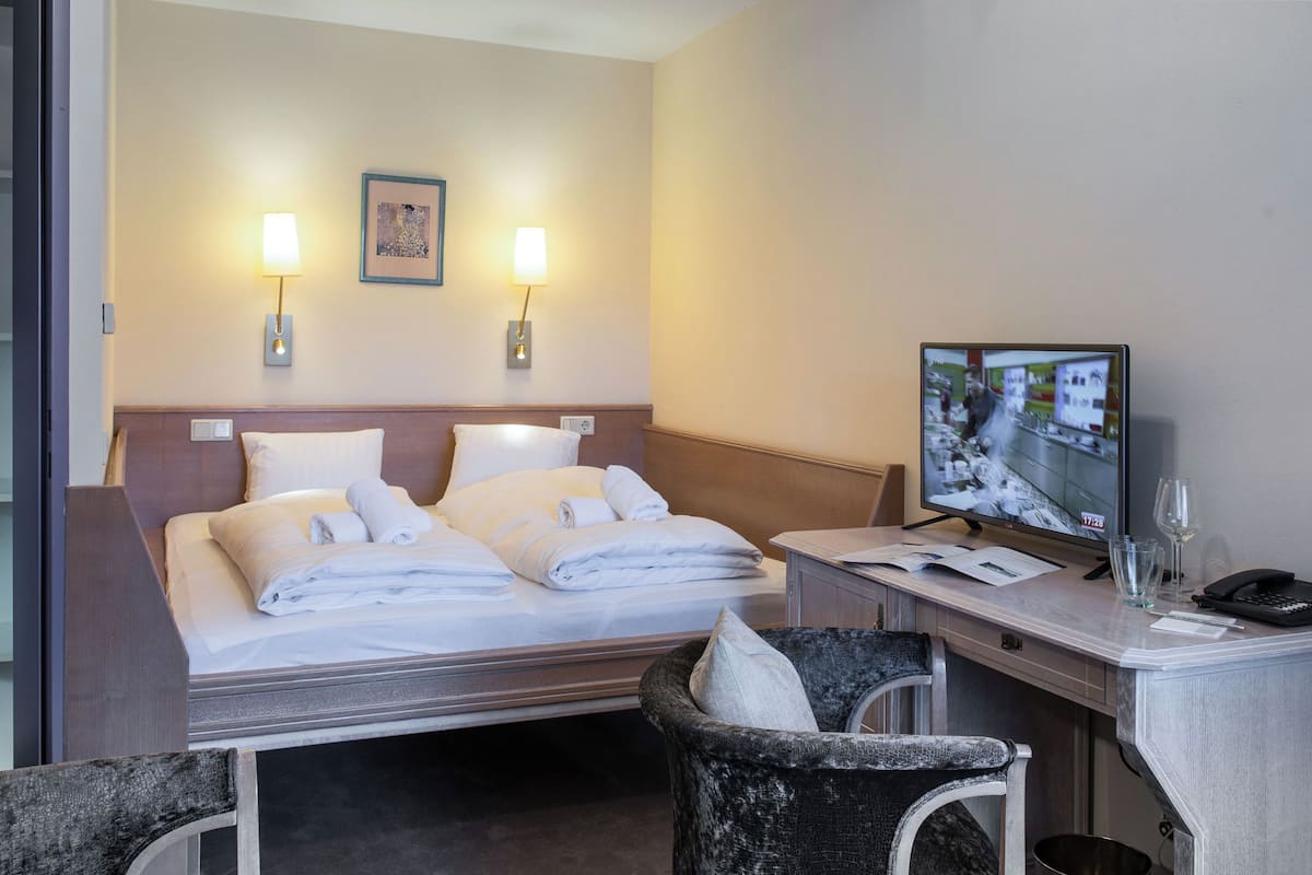 Quarto de hotel com cama, secretária, cadeira, televisão e duas luminárias fixadas na parede.