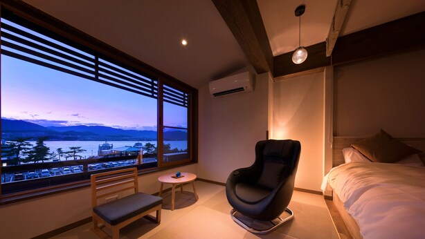 Quarto de hotel com uma janela grande, uma cama, uma cadeira, uma mesa pequena e um aparelho de ar condicionado.